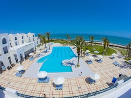 Uma área de piscina em resort, com vista clara do oceano, espreguiçadeiras, guarda-sóis e um edifício branco com arcos.