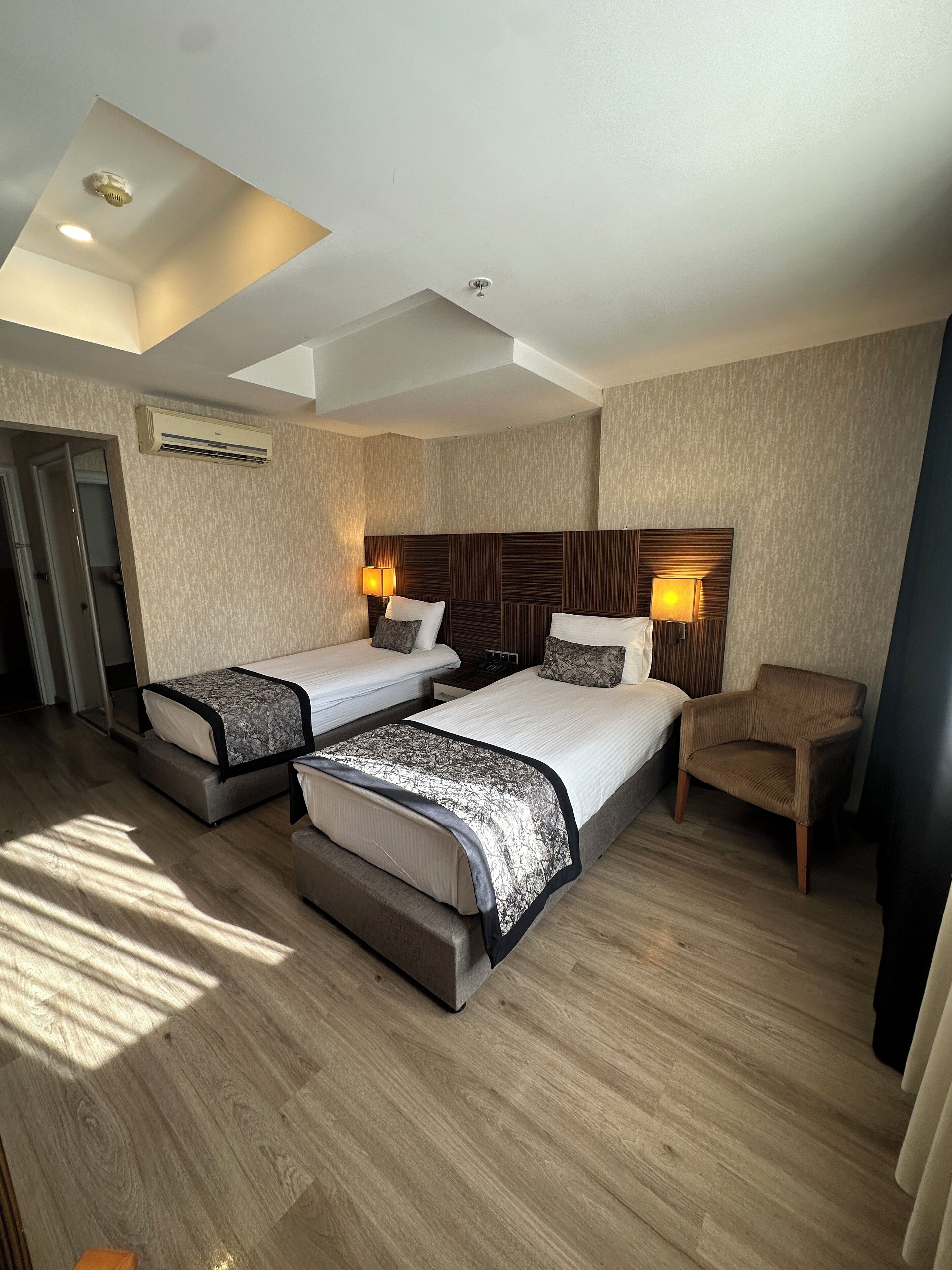 Quarto de hotel com duas camas, piso de madeira, uma cadeira e uma luminária no teto.