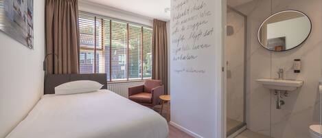 Quarto de hotel com uma cama, uma cadeira, uma mesa pequena, banheiro com pia e espelho, e uma parede com texto.
