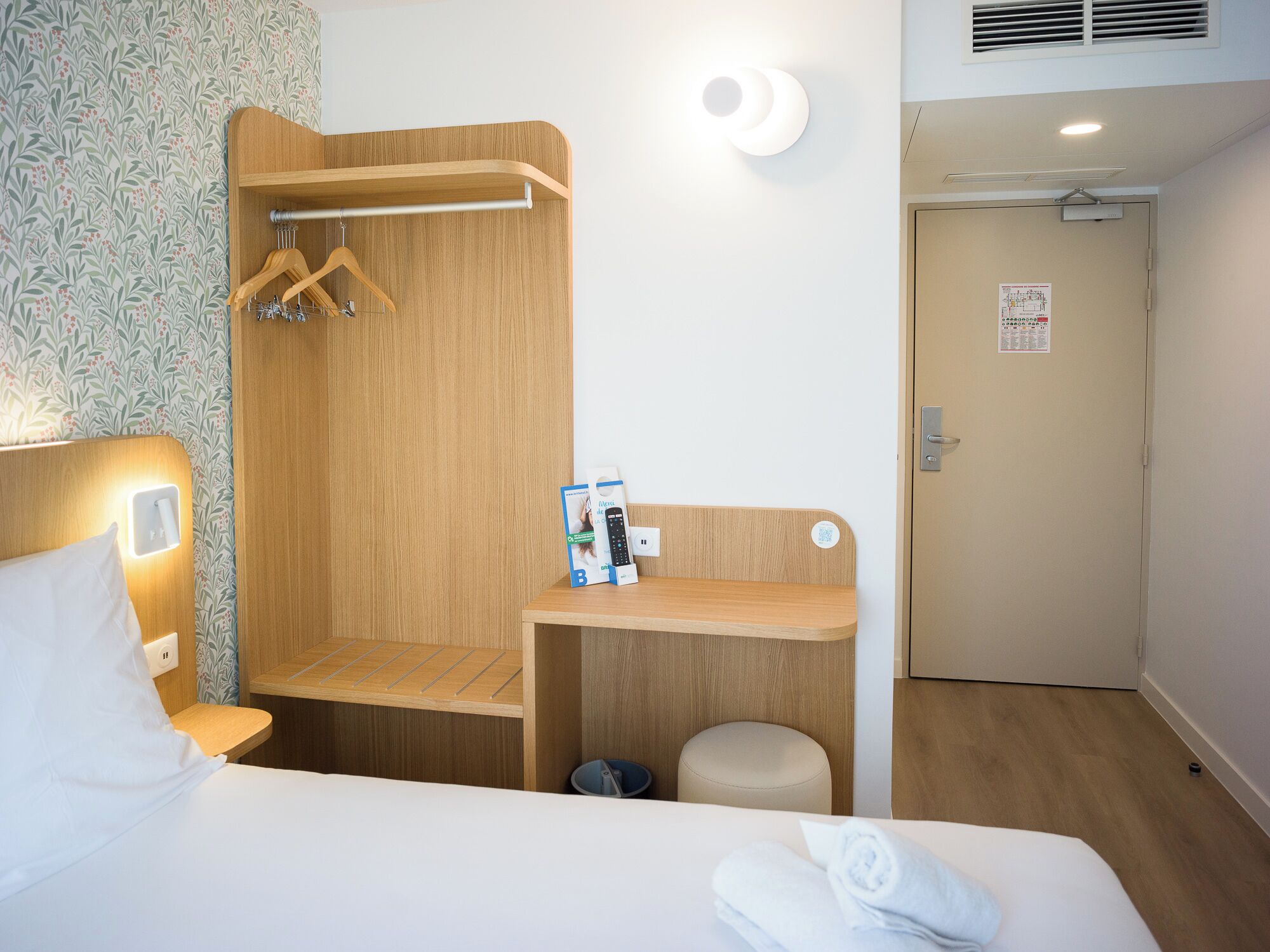 Quarto de hotel com cama, área de chuveiro em madeira, um pequeno compartimento com um controle remoto e porta com sinal de segurança.