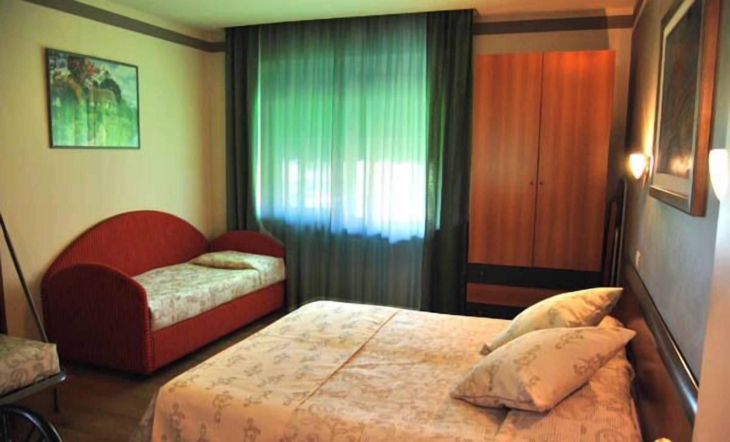 Quarto de hotel com cama, sofá vermelho, quadro na parede e janela com cortinas.