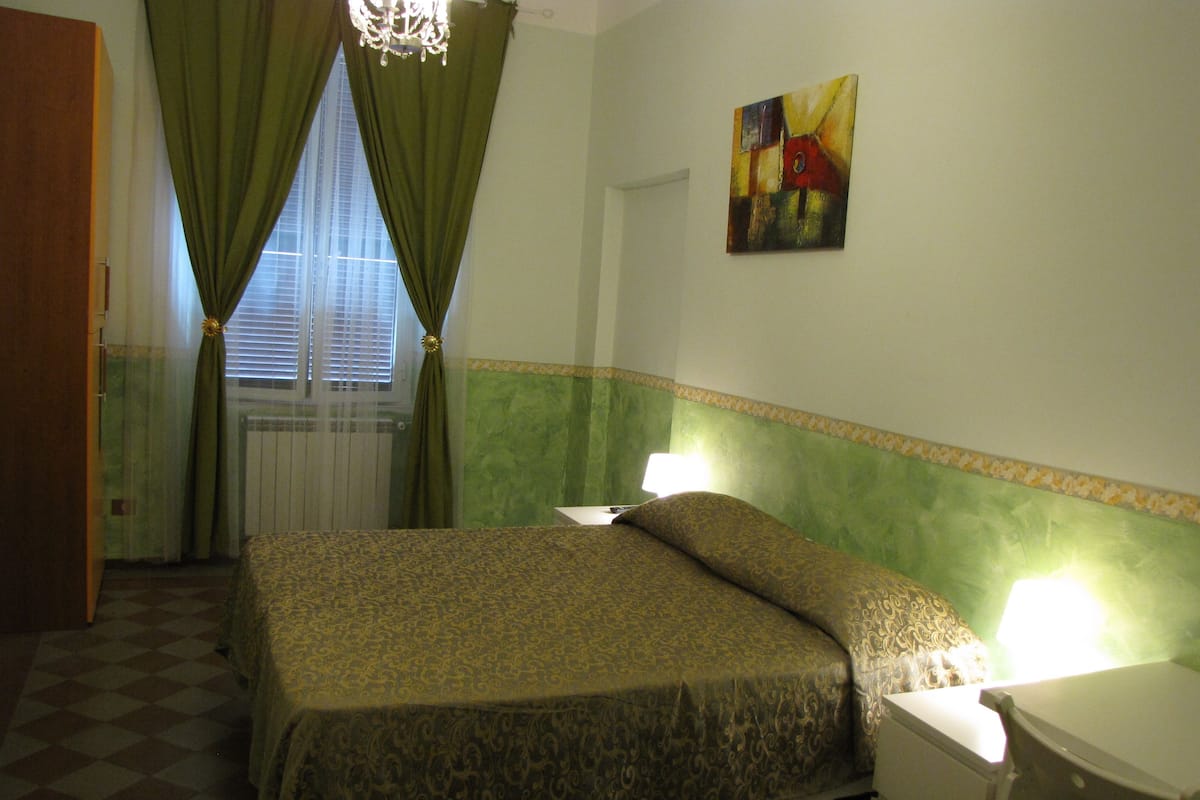 Um quarto com uma cama, mesa de cabeceira, luminária, janela com cortinas e um quadro na parede.