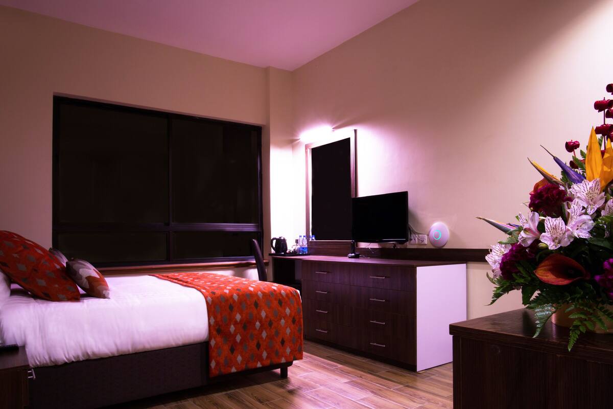 Quarto com cama, penteadeira, televisão e uma janela grande.