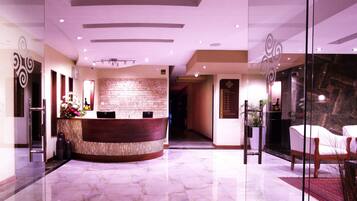 Uma área de recepção de hotel com piso de mármore, balcão de recepção, parede de destaque em tijolo e um espaço de estar com cadeiras brancas.