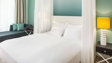 Quarto de hotel moderno com uma cama grande, mesa de cabeceira com abajur, sofá com almofadas e cortinas leves.