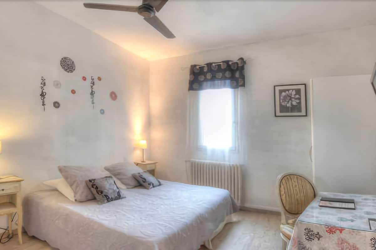 Quarto com cama, mesas de cabeceira, poltrona, mesa, ventoinha no teto, janela com cortinas, radiador e decorações nas paredes.