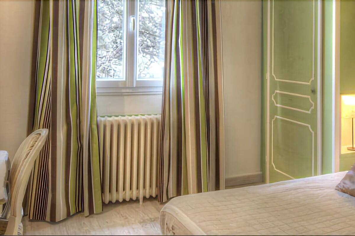 Quarto com cama, almofadas, radiador, cortinas e parede com um desenho floral.