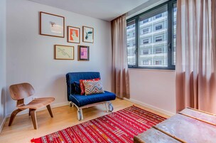 Uma sala de estar moderna com um sofá azul, uma cadeira de madeira, um tapete vermelho estampado e obras de arte emolduradas na parede.