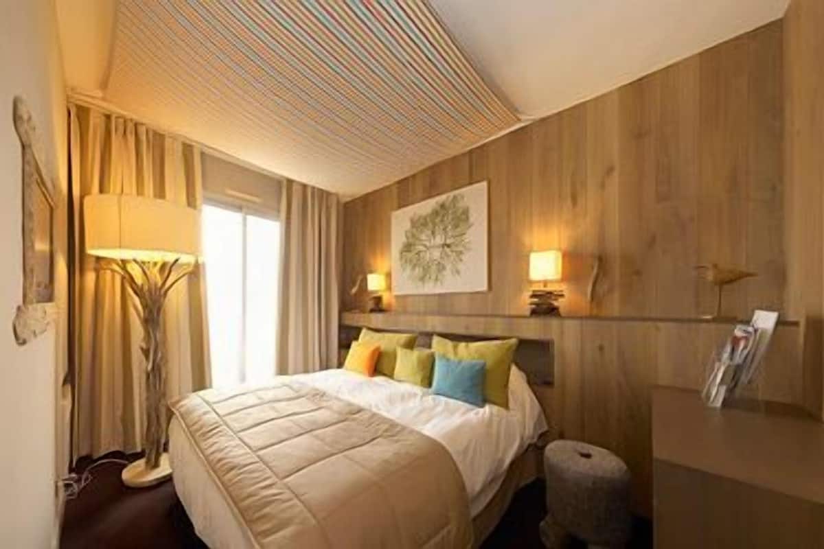 Quarto de hotel com uma cama grande, um abajur, uma cabeceira de madeira e uma prateleira fixada na parede com uma revista.