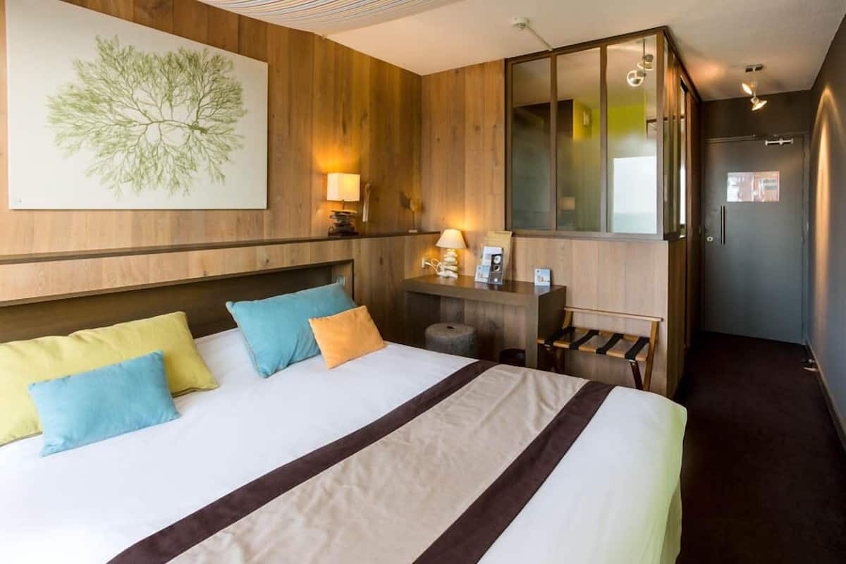 Quarto de hotel com uma cama grande, cabeceira de madeira, mesa de cabeceira com luminária, espelho e vista para o corredor.