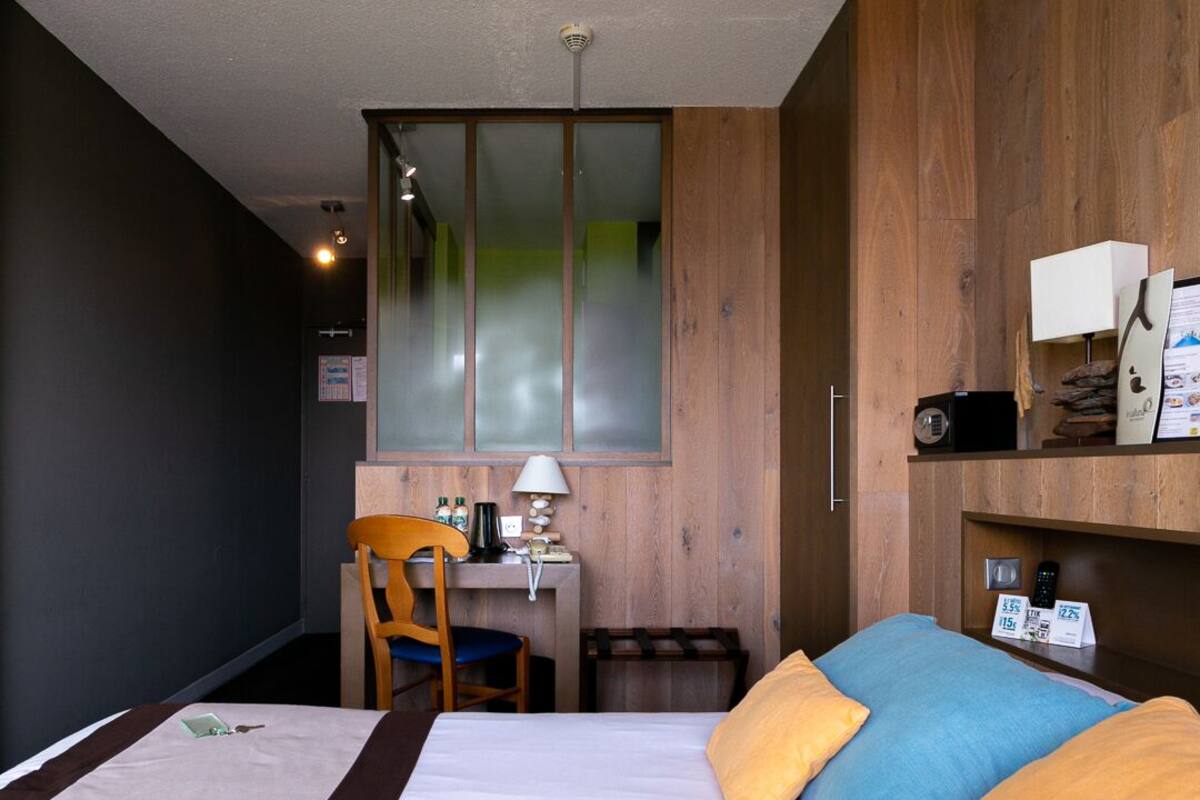 Quarto de hotel com cama, escrivaninha, cadeira e pequena mesa com abajur.