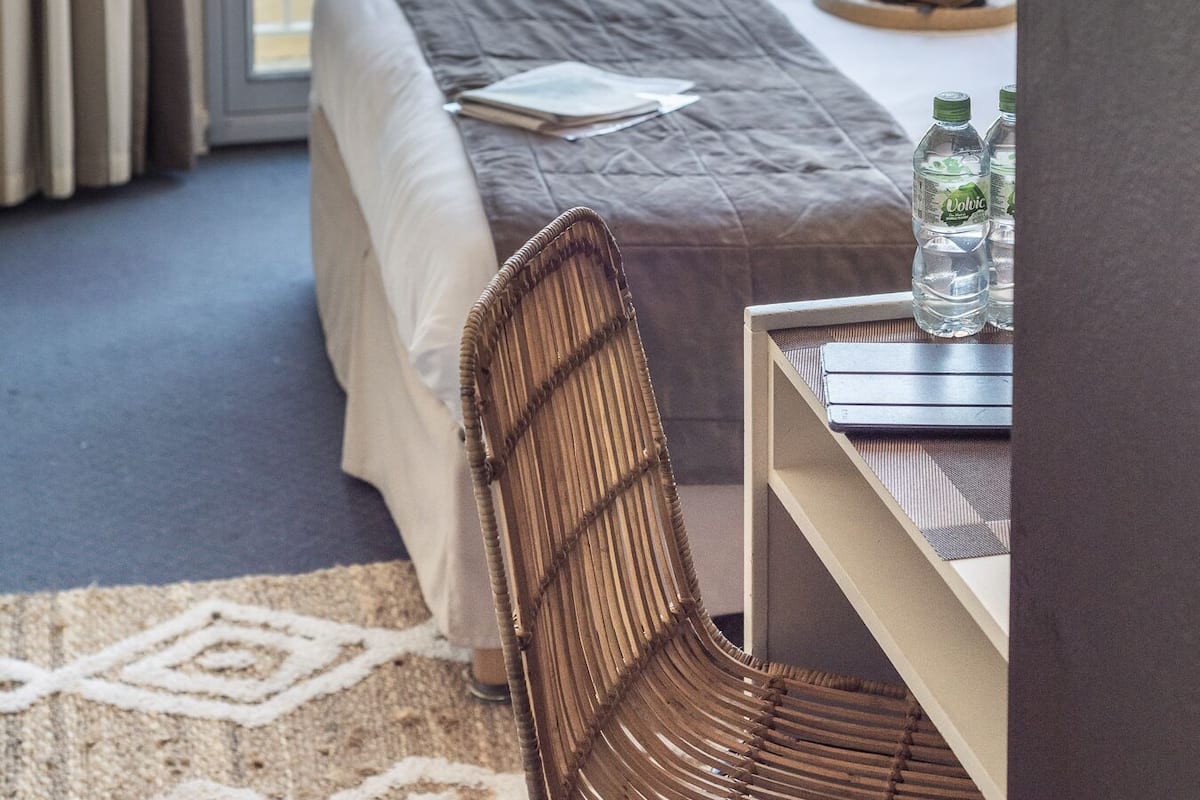 Quarto de hotel com cama, uma cadeira, uma mesa com garrafas de água e vista para o mar com barcos.