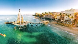 Um hotel à beira-mar com uma ponte que se estende até o oceano, comodidades na orla e montanhas ao fundo.