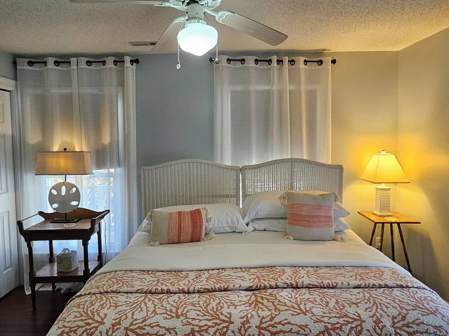 Quarto com uma cama, duas mesas de cabeceira com luminárias, um ventilador de teto e uma janela com cortinas.