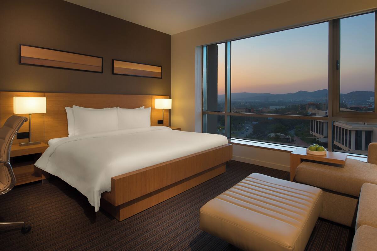 Quarto de hotel moderno com uma cama grande, duas luminárias de cabeceira, uma poltrona, uma mesinha com uma cesta de frutas e uma grande janela com vista para a cidade.