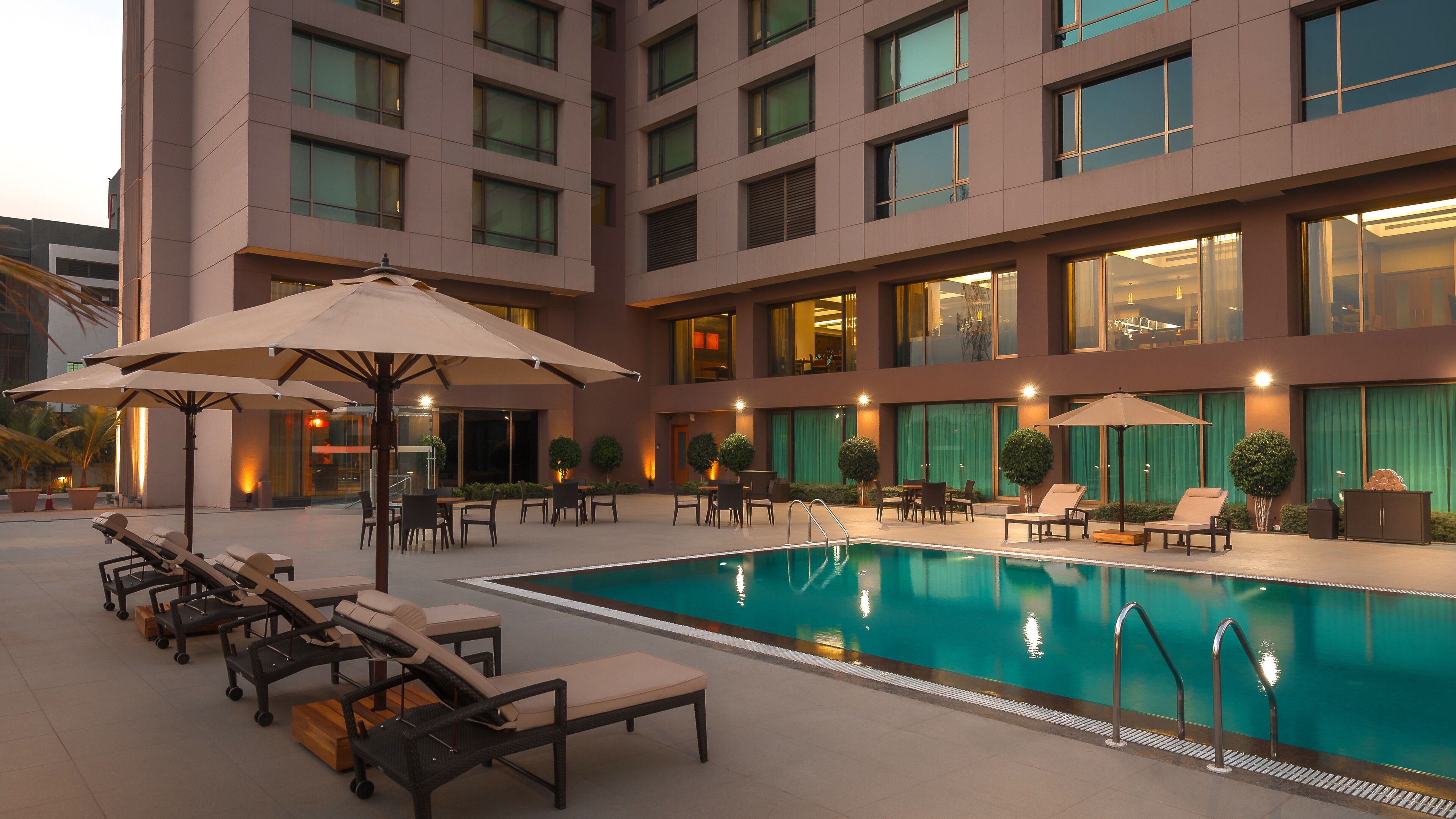 Área de piscina de hotel com espreguiçadeiras e guarda-sóis, cercada por um edifício com vários andares e grandes janelas.