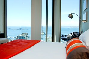 Quarto de hotel com uma grande janela que oferece vista para o mar e para a paisagem urbana ao longe.