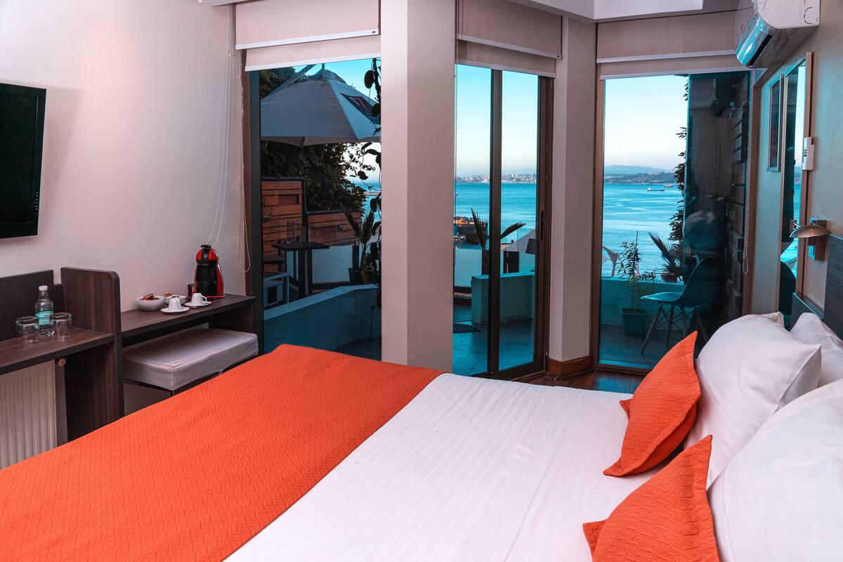 Quarto de hotel com uma cama grande, roupa de cama laranja, uma mesa com cadeira e vista para o mar através de uma porta de vidro.