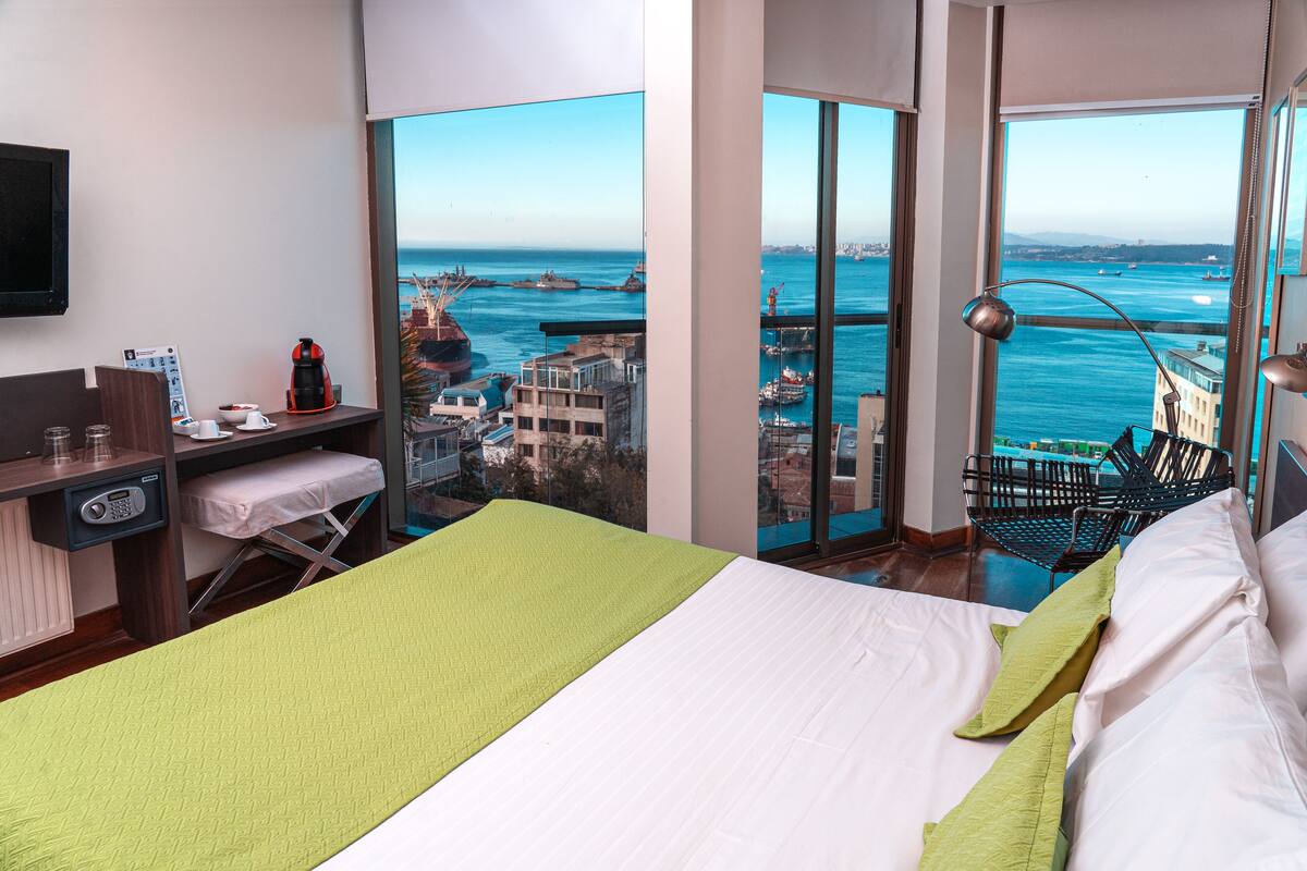 Quarto de hotel com uma cama grande, uma escrivaninha, uma cadeira e vista para o mar e a paisagem urbana.