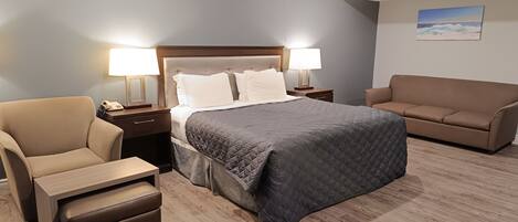 Quarto de hotel com uma cama, duas luminárias de cabeceira, uma poltrona de couro marrom, um sofá marrom e piso de madeira.