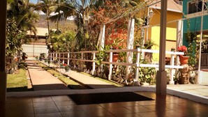 Uma trilha pavimentada conduz a um portão com cerca branca, em meio a vasos de plantas e diversas árvores.