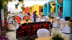 Uma área de refeições ao ar livre com mesas e cadeiras, algumas com toalhas de mesa brancas e outras amarelas.