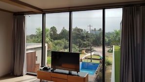 Uma sala de estar moderna com uma televisão grande sobre um armário de madeira, janelas amplas com cortinas e vista para uma piscina e vegetação lá fora.