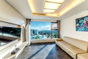 Uma sala de estar moderna com uma televisão grande, um sofá, um quadro e vista para uma piscina e edifícios.
