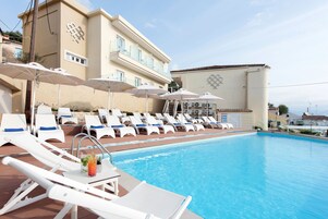 Piscina com espreguiçadeiras brancas e guarda-sóis, um deck revestido de azulejos e um edifício com varandas e janelas.