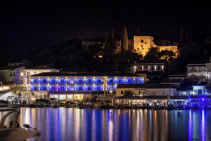 Um hotel à beira-mar com um castelo imponente ao fundo, uma marina com barcos e um edifício com varandas iluminadas em azul.
