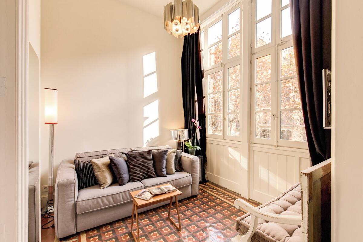 Sala de estar com sofá, mesa de centro e uma poltrona. Há uma janela com cortinas, uma luminária e uma planta.
