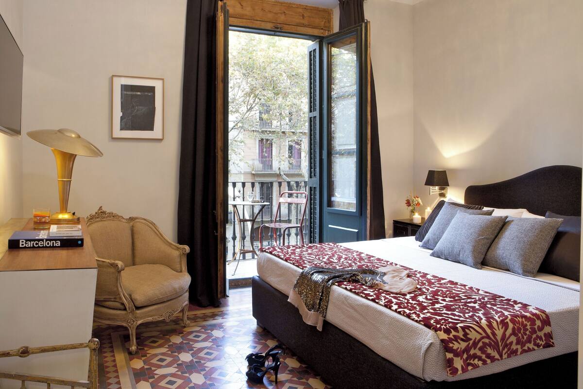 Quarto de hotel com uma cama, uma cadeira, uma escrivaninha com luminária e vista para a varanda.