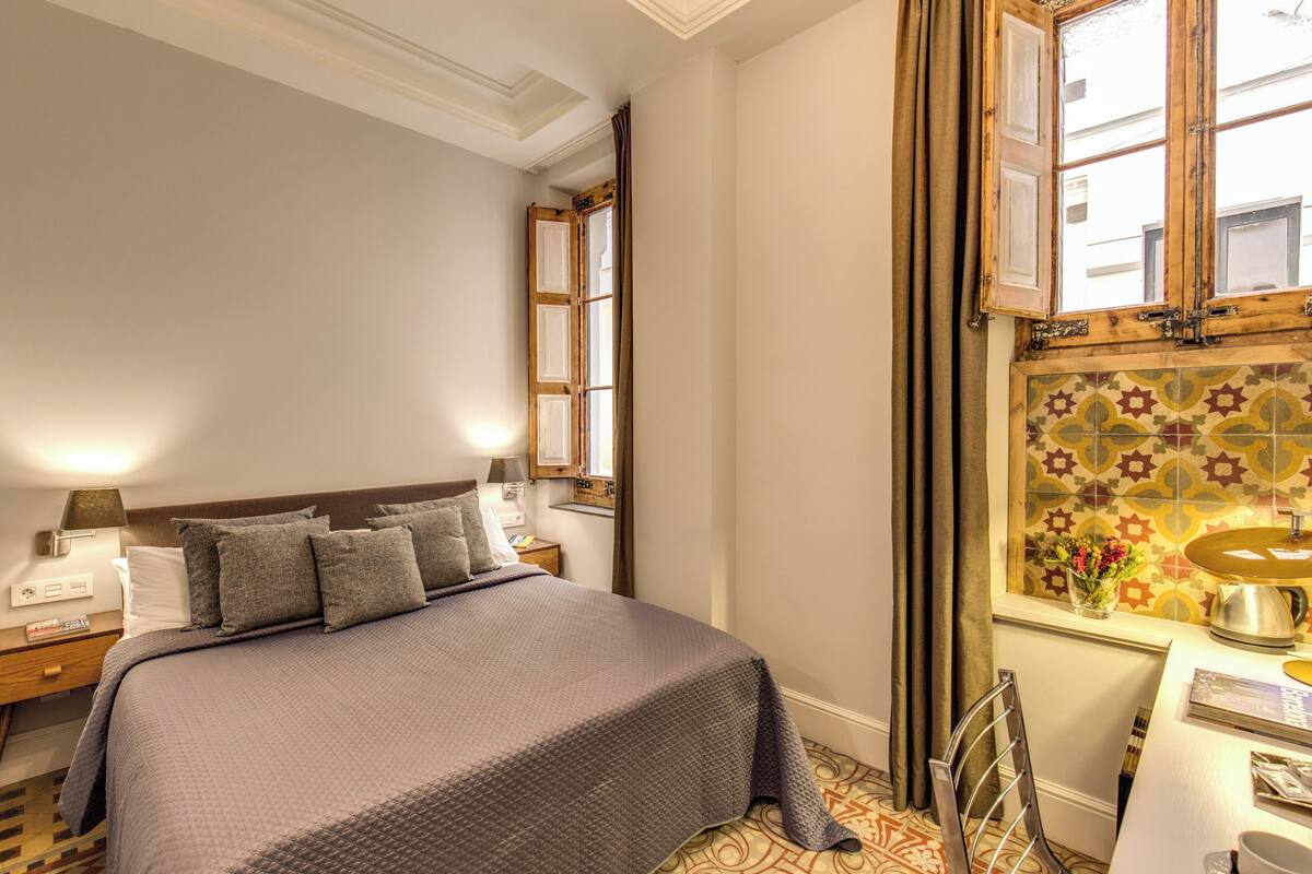 Quarto de hotel com cama, mesa de cabeceira, abajur e janela com cortinas.