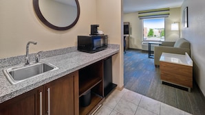 Quarto de hotel com bancada de granito, pia, micro-ondas, cafeteira, espelho, pequeno espaço de estar com uma cadeira e mesa, e janela com vista.