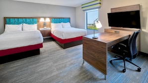 Quarto de hotel com duas camas, uma escrivaninha com televisão, uma cadeira, uma janela com vista e uma mesa de cabeceira com abajur.