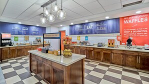 Espaço de café da manhã com estação de café, variedade de frutas e equipamentos para waffles.
