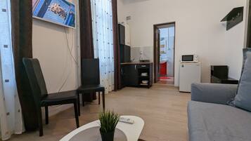 Uma sala de estar moderna com um sofá cinzento, uma poltrona preta, uma pequena mesa com uma planta, uma TV fixada na parede e uma área de cozinha visível através de uma porta aberta.