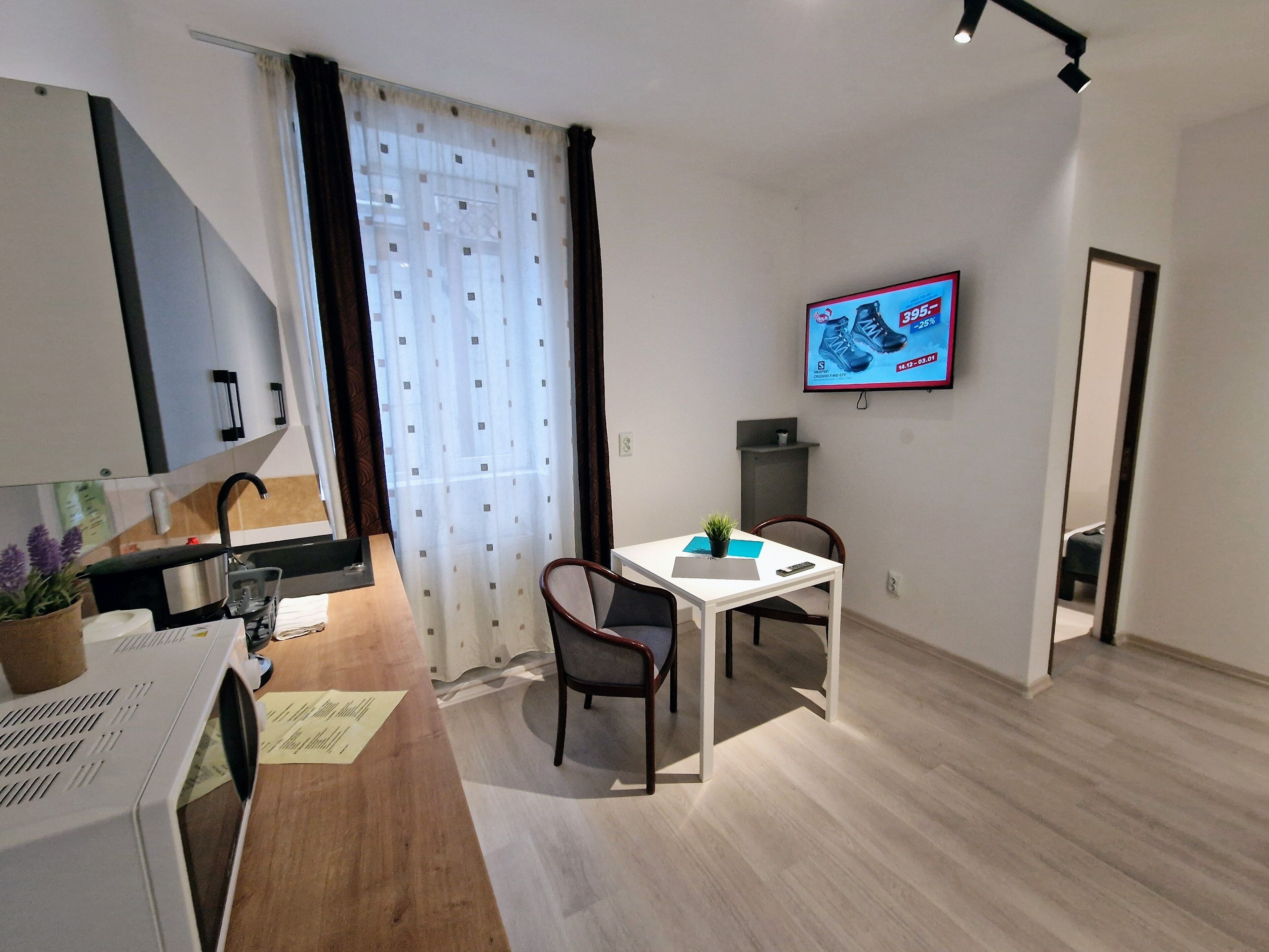 Quarto moderno com mesa de jantar, cadeiras, TV na parede e área de cozinha com micro-ondas e chaleira.