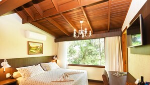Um quarto com cama, mesa de cabeceira, escrivaninha com cadeira, lustre, janela com cortinas e um quadro na parede.