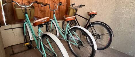 Duas bicicletas estacionadas em ambiente interno, uma com estrutura azul-turquesa e outra com estrutura marrom, encostadas a uma parede de madeira.