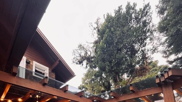 Uma área de estar ao ar livre coberta, com pérgola de madeira, uma bicicleta e diversos vasos com plantas e flores.
