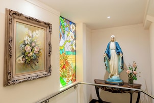 Uma estátua de figura religiosa sobre um pedestal, um quadro emoldurado de flores e uma janela de vitrais.