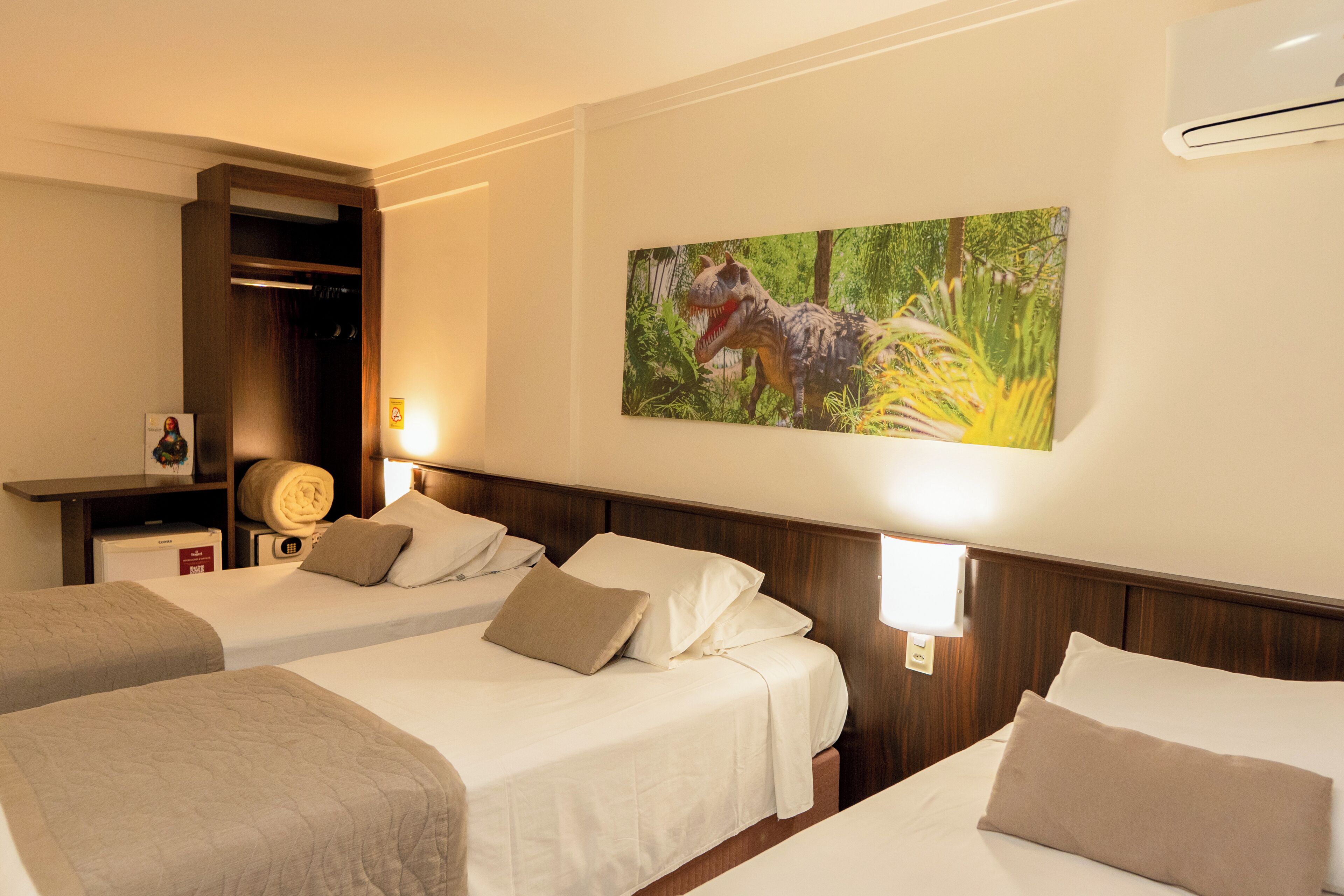 Quarto de hotel com duas camas, cabeceira de madeira, pintura de um dinossauro e uma pequena estante com uma toalha enrolada.