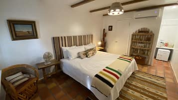 Quarto com cama grande, cabeceira de madeira, mesa de cabeceira com ventilador, cadeira de balanço de madeira, tapete estampado e uma pequena cozinha compacta com geladeira.