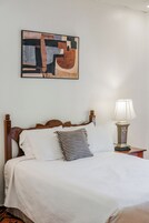 Cama bem arrumada com edredom branco e travesseiro listrado, cabeceira de madeira, abajur e pintura abstrata na parede.