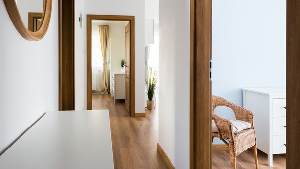 Um corredor com piso de madeira, uma escrivaninha branca, uma cadeira de vime e um vaso com planta.