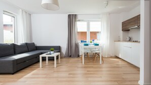 Uma sala de estar moderna com um sofá cinzento, uma pequena mesa branca com uma planta, e uma área de jantar com uma mesa e cadeiras brancas.
