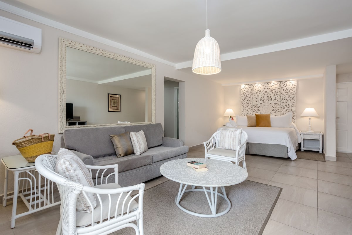 Quarto de hotel moderno com sofá cinzento, cadeiras de vime branco, mesa redonda, espelho grande e cama com roupa de cama branca.