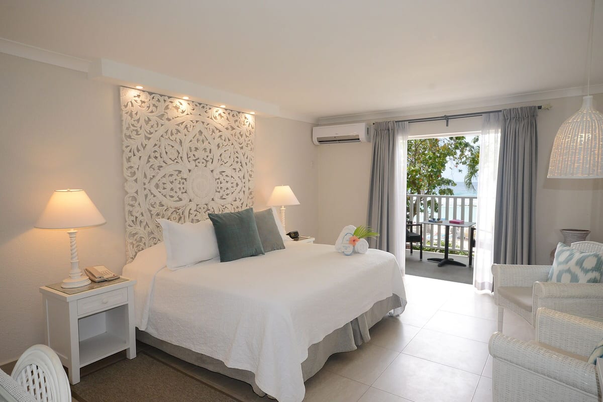 Quarto de hotel com uma cama grande, duas mesas de cabeceira, um criado-mudo, uma cadeira, uma varanda com mesa e cadeiras, e um lustre.