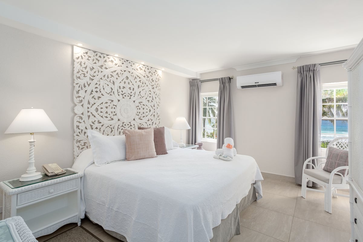 Quarto de hotel com uma cama grande, duas mesas de cabeceira com abajures, uma cabeceira decorativa, uma janela com cortinas e vista para o oceano.